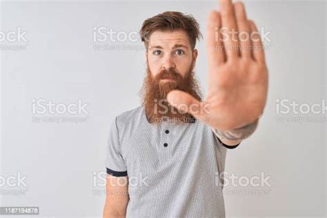 고립 된 흰색 배경 위에 서있는 회색 폴로를 입은 젊은 빨간 머리 아일랜드 남자가 손바닥으로 노래를 멈추고 있습니다 얼굴에