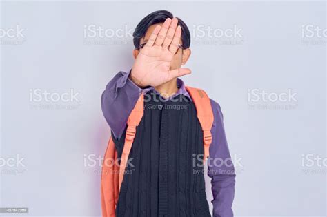 배낭을 메고 캐주얼한 옷을 입은 진지한 젊은 아시아 학생 남자가 회색 배경에 고립된 열린 손바닥으로 얼굴을 가리고 제스처를