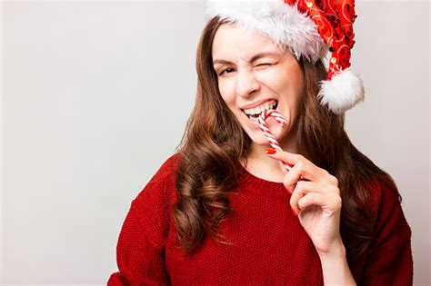 빨간 스웨터와 산타 모자에 쾌활한 재미 젊은 여자는 줄무늬 롤리팝을 물고있다 공휴일에 대한 스톡 사진 및 기타 이미지 공휴일 이빨 긍정적인 감정 표현 Istock