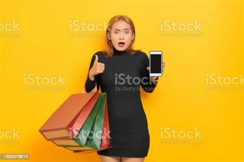 깜짝 놀란 젊은 아시아 여자 빈 화면 휴대 전화를 들고 엄지 손가락을 몸짓 노란색 배경에 고립 된 쇼핑 백을 들고 25 29세에