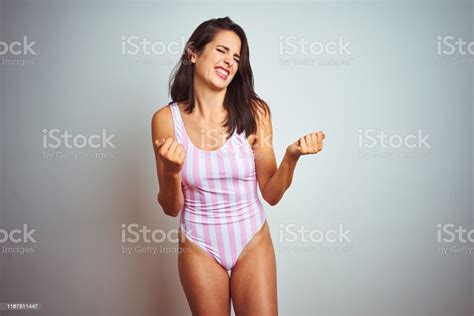 고립 된 배경에 줄무늬 핑크 수영복 수영복을 입은 젊은 아름다운 여성은 매우 행복하고 팔을 들고 미소를 지으며 성공을 위해