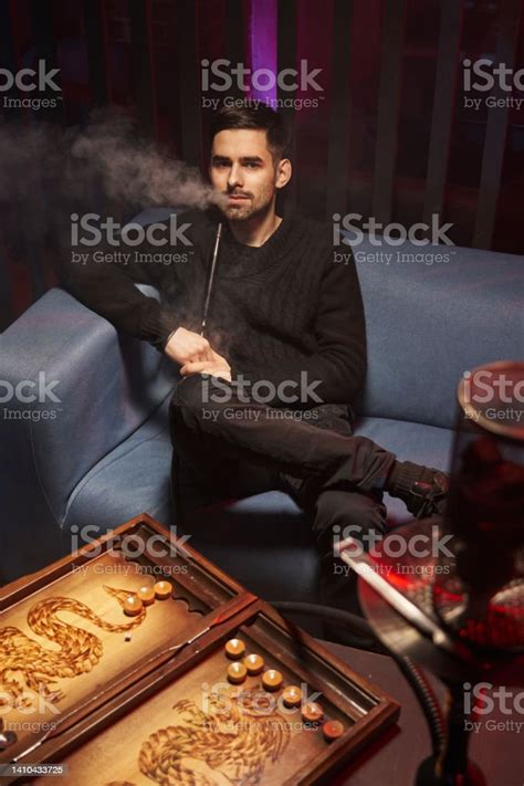 한 젊은이가 컬러 필터로 조명 된 대기 담뱃대 바에서 물 담뱃대를 피우고 손에 담뱃대 파이프를 들고 하얀 연기를 내뿜습니다 25