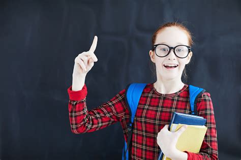 명랑 한 흥분 한 여 학생 안경에 체크 무늬 드레스와 가방을 착용 하는 동안 아이디어를가지고 그녀는 칠판에 서 서 공부에 대한