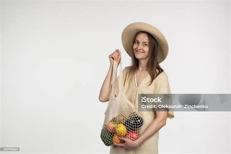 가벼운 드레스를 입고 챙이 넓은 밀짚모자를 쓴 여성이 형형색색의 과일이 가득 담긴 그물 가방을 들고 미소를 짓고 있다 그녀는 평범한 배경에 서 있다 30 34세에 대한 스톡