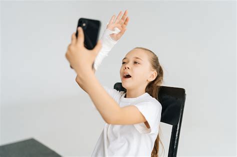 흰색 석고 붕대로 싸인 부러진 팔을 가진 귀여운 금발 소녀가 흰색 고립된 배경에 의자에 앉아 스마트폰으로 화상 통화를 하고 있다 8 9 살에 대한 스톡 사진 및 기타 이미지