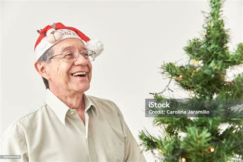 빨간 산타 모자를 쓰고 크리스마스 트리 옆에 미소 짓는 행복한 노인 라틴계 남자 12월에 대한 스톡 사진 및 기타 이미지 12월 공휴일 긍정적인 감정 표현 Istock