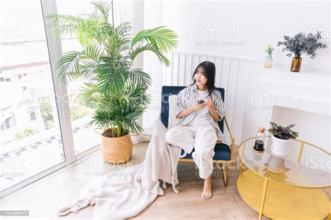 젊은 예쁜 여자는 미소를 짓고 녹색 식물이있는 현대 거실의 의자에 앉아 뜨거운 커피 한 잔을 마시 며 휴식을 취하고 아늑한