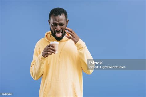 파란색 배경에 고립 된 일회용 커피 컵을 들고 얼굴에 얼굴을 찡 그리기 함께 젊은 아프리카계 미국인 남자를 화나게 감정에 대한