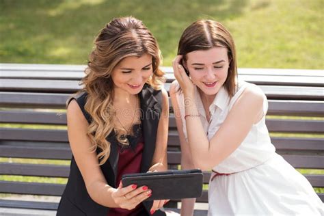 Two Young Girls Sitting On A Bench In A Park Watching The Tablet And