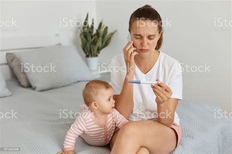 아기 딸과 함께 침대에 앉아 전화를 말하는 흰색 티셔츠를 입고 임신 검사를 손에 들고 부정적인 감정을 표현하는 슬픈 화가 여성의