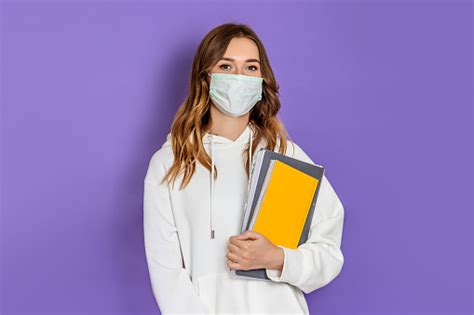 수술용 의료용 마스크를 쓴 어린 갈색 머리 학생 소녀가 하얀 후드티를 입고 서서 라일락 배경에 고립된 카메라를 바라보는 모습 30 34세에 대한 스톡 사진 및 기타 이미지