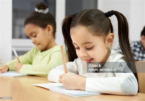 테이블에 앉아서 카피 북에 쓰는 매력적인 행복 한 학생의 측면도 작은 여성 학생 펜을 유지 하 고 그림 학습과 학교에서 공부 지식의 개념입니다 우정에 대한 스톡 사진 및