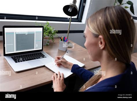 Caucasian Female Programmer Sitting At Desk Using Laptop With Coding On Screen Making Notes
