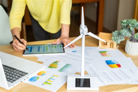 여성 사업가는 재생 에너지에 관한 프로젝트를 진행하고 있으며 책상 위에 놓인 소형 풍력 터빈과 태양 전지판을 차트와 그래프와 함께 디지털 태블릿의 데이터를 분석하고 있습니다