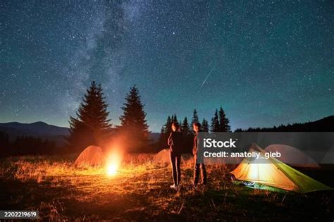두 명의 등산객이 별이 총총한 광활한 하늘 아래 모닥불 옆에 서서 배경으로 은은하게 빛나는 텐트 근처에 서 있습니다 2명에 대한 스톡 사진 및 기타 이미지 Istock