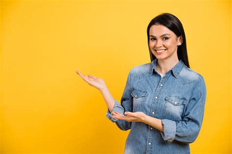 유쾌한 젊은 갈색 머리 여자 캐주얼 세련 된 파란색 셔츠를 입고 친절한 미소와 측면에 손을 가리키는 고립 된 오렌지 배경에 서