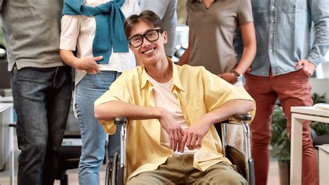 나는 여기에서 일하게되어 너무 행복하다 휠체어를 탄 젊고 긍정적인 아시아 남자의 초상화가 카메라를 바라보며 미소짓고 있는 동안 동료들이 배경에 서 있는 모습 169에 대한