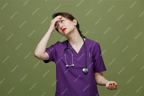 首の周りに制服と聴診器を身に着けている痛む中年の女性医師は、オリーブグリーンの背景に頭痛が分離されている側を見て頭に手を保ちます 無料の写真
