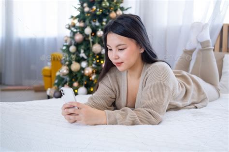 Happy Beautiful Asian Girl In Cozy Clothing Using Phone Lying On Bed At