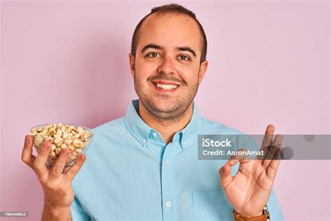 피스타치오가 서있는 그릇을 들고있는 젊은 남자는 손가락으로 확인 기호를하고 우수한 기호를하고 고립 된 분홍색 배경 위에 서 남자에 대한 스톡 사진 및 기타 이미지 Istock