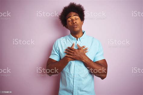 고립된 분홍색 배경 위에 서있는 파란 셔츠를 입은 젊은 미국 남자는 닫힌 눈과 얼굴에 감사하는 제스처로 가슴에 손을 미소를 짓고 있습니다 건강 개념 감사에 대한 스톡 사진