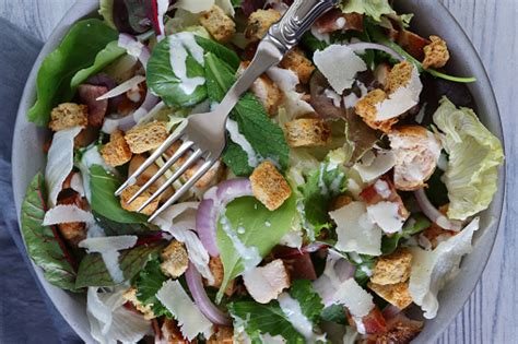 그릇에 치킨 시저 샐러드의 클로즈업 이미지 양상추 붉은 양파 비트 뿌리 잎 큐브 베이컨 구운 닭고기 크루통과 파마산 치즈 면도 회색 차 수건 외에 금속 포크 나무 곡물 배경