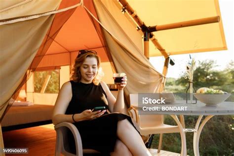 행복한 여성 프리랜서는 화창한 날에 아늑한 글램핑 텐트에서 휴식을 취하면서 전화와 칵테일 통화를 사용합니다 야외 여름 휴가 및 휴가를 위한 고급 캠핑 텐트 라이프스타일 컨셉
