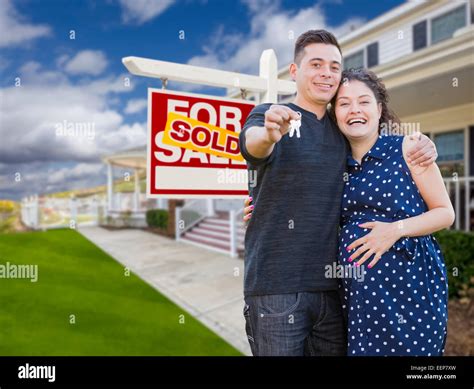 Happy Hispanic Couple In Front Of New Home And Sold Real Estate Sign