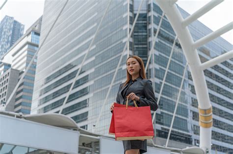 회색 사무실 정장을 입은 젊은 아시아 사업가가 빨간 쇼핑 종이 봉지를 손에 들고 중앙 비즈니스 지구를 행복하게 걷고 있다 18 19세에 대한 스톡 사진 및 기타 이미지