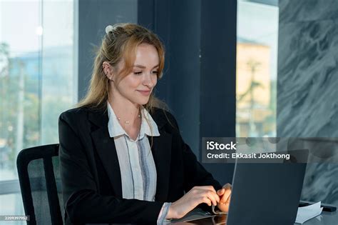 비즈니스 정장을 입은 젊은 백인 여성이 사무실 책상에서 노트북으로 일하면서 큰 유리창을 통해 들어오는 자연 채광이 들어오는 조용한 기업 환경에서 자신 있게 미소 짓고