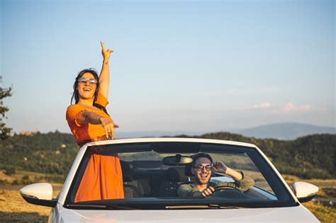 이동 중에도 두 명의 연인을위한 일몰 일광욕 여행 및 휴가 라이프 스타일 컨버터블 자동차 시골 배경에 서서 즐거운 시간을 보내는 젊은 커플 팔 들기에 대한 스톡 사진 및