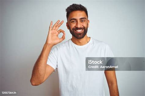 고립 된 흰색 배경 위에 서있는 티셔츠를 입은 젊은 인도 남자가 손과 손가락으로 확인 기호를하고 긍정적 인 미소를 짓고 있습니다 성공적인 식입니다 남자에 대한 스톡 사진 및
