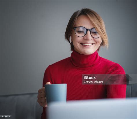 빨간 스웨터와 안경을 쓴 어른의 아름다운 여성이 노트북 화면 앞에서 손에 머그잔을 들고 소파에 앉아 있는 모습을 볼 수 있습니다