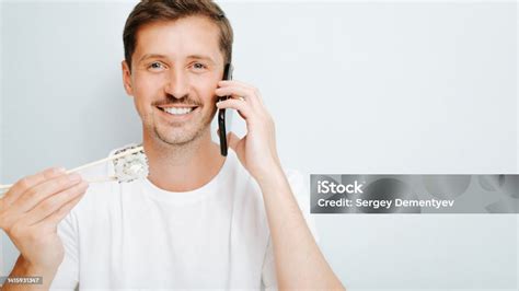 초밥을 먹고 파란 배경에 서서 카메라를 바라 보는 휴대 전화를 사용하는 긍정적 인 남자의 초상화 젓가락으로 롤을 들고 스마트 폰으로 전화하는 웃는 남자 복사 공간 30 34