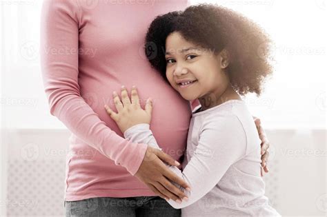 Close up of little black daughter hugging her pregnant mother's tummy, standing next to window ...
