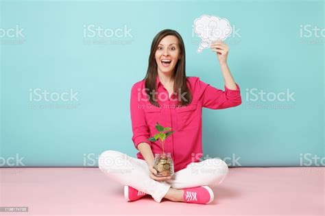 장미 셔츠에 여자의 전체 길이 흰색 바지 바닥에 앉아 밝은 분홍색 파란 파스텔 벽 배경 스튜디오에 고립 된 유리 항아리에 금화