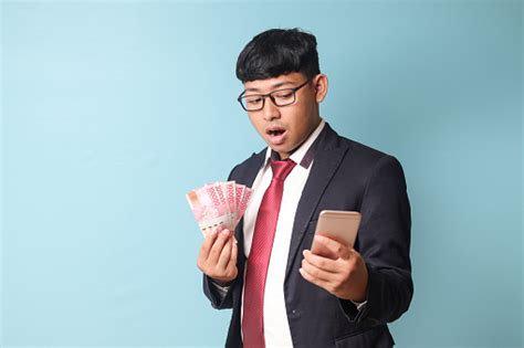 캐주얼 정장을 입은 젊은 아시아 사업가의 초상화는 수천 루피아를 들고 놀란 스마트폰을 보고 있다 파란색 배경에 격리된 이미지
