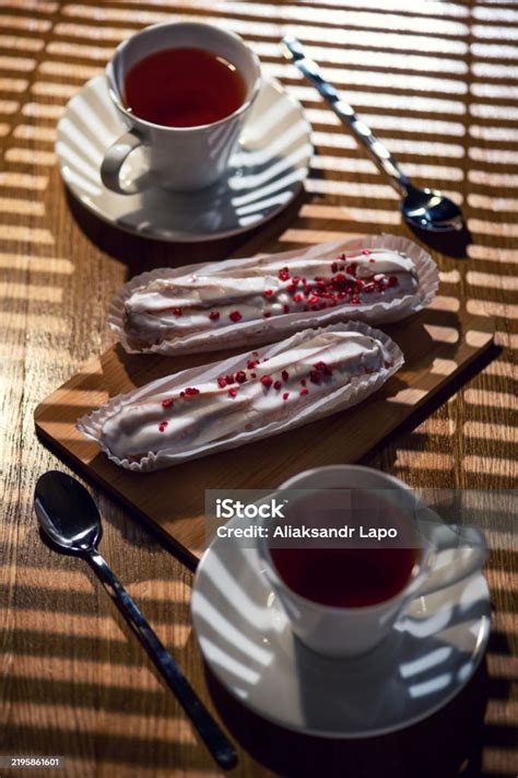 접시에 담긴 차 두 잔과 숟가락이 놓여 있고 나무 판자에 하얀 아이싱과 빨간 스프링클을 얹은 에클레어 두 잔이 줄무늬 그림자 패턴이 있는 테이블 위에 놓여 있습니다 0명에