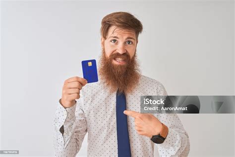 자신에게 손가락을 가리키는 깜짝 얼굴과 고립 된 흰색 배경 위에 서있는 신용 카드를 들고 젊은 빨간 머리 아일랜드어 사업가 신용 카드에 대한 스톡 사진 및 기타 이미지