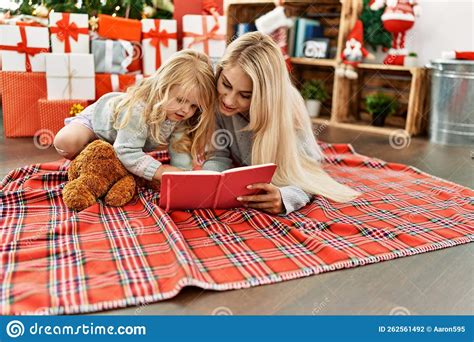 Mother And Daughter Reading Book Lying By Christmas Tree At Home Stock