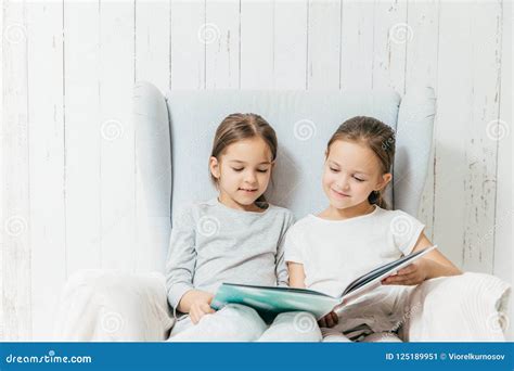 Two Little Adorable Sisters Sit In Sofa Read Interesting Book Stock