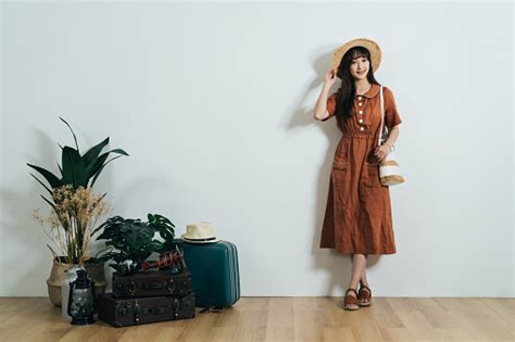 여름 드레스에 다리를 꼬고 서있는 전체 길이의 아시아 여성은 고립 된 흰색 벽 배경에 대해 카메라를 미소 짓는 동안 밀짚 모자를 들고 있습니다 개념에 대한 스톡 사진 및