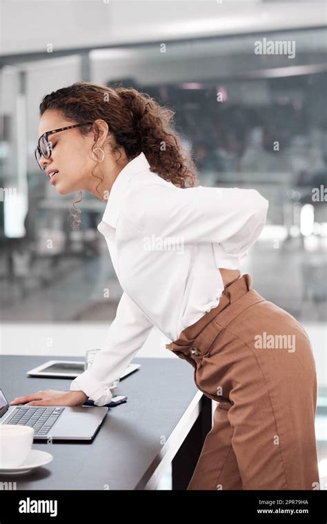 A Woman Holding Her Back In Pain And Leaning Against Her Desk Stock