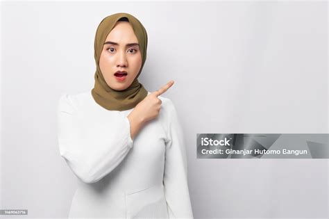 히잡을 쓰고 흰색 배경에 격리된 빈 복사 공간을 손가락으로 가리키며 서 있는 아름다운 충격을 받은 젊은 아라비아 아시아 무슬림