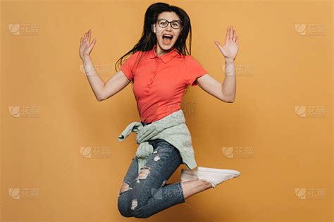 年轻的黑发女孩戴着眼镜，身穿红色t恤、牛仔裤，腰上系着毛衣，脚上穿着白色运动鞋，在黄色的背景下，蹦蹦