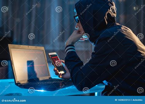 Male Hacker In The Hood Holding The Phone In His Hands Trying To Steal Access Databases With