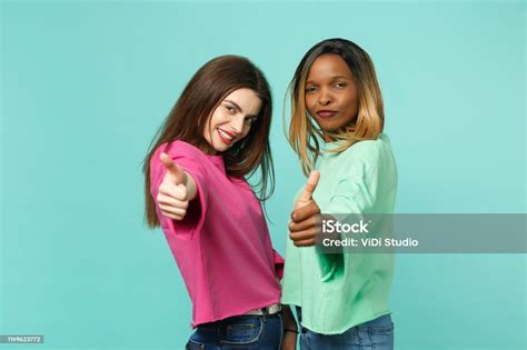 두 젊은 여자 친구 파란색 청록색 벽 배경 스튜디오 초상화에 고립 된 엄지손가락을 보여주는 분홍색 녹색 옷에 유럽과 아프리카계