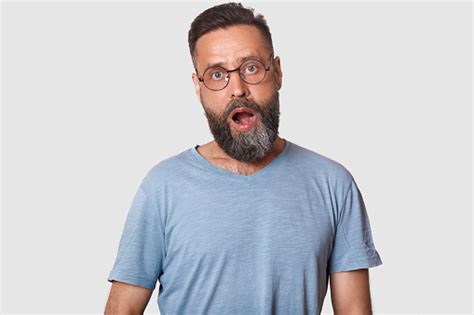 수염을 기운 백인 남자의 실내 샷은 예기치 않은 무언가를 통지 널리 입을 열어 유지 놀란되고 남성은 흰색 콘크리트 벽에 고립 된