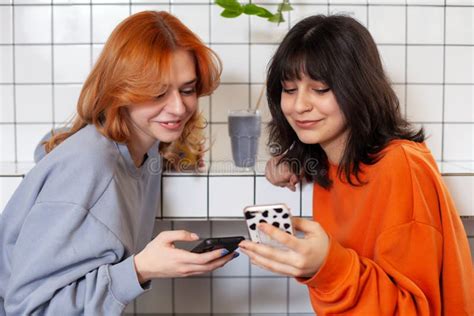 Two Excited Young Girls Using Mobile Phones While Sitting At The Cafe And Pointing Finger Stock