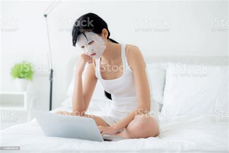 침실에서 침대에 노트북 컴퓨터에 앉아 시트 얼굴 마스크를 가진 아름다운 젊은 아시아 여성 집에서 주름의 스킨 케어에 대한 얼굴